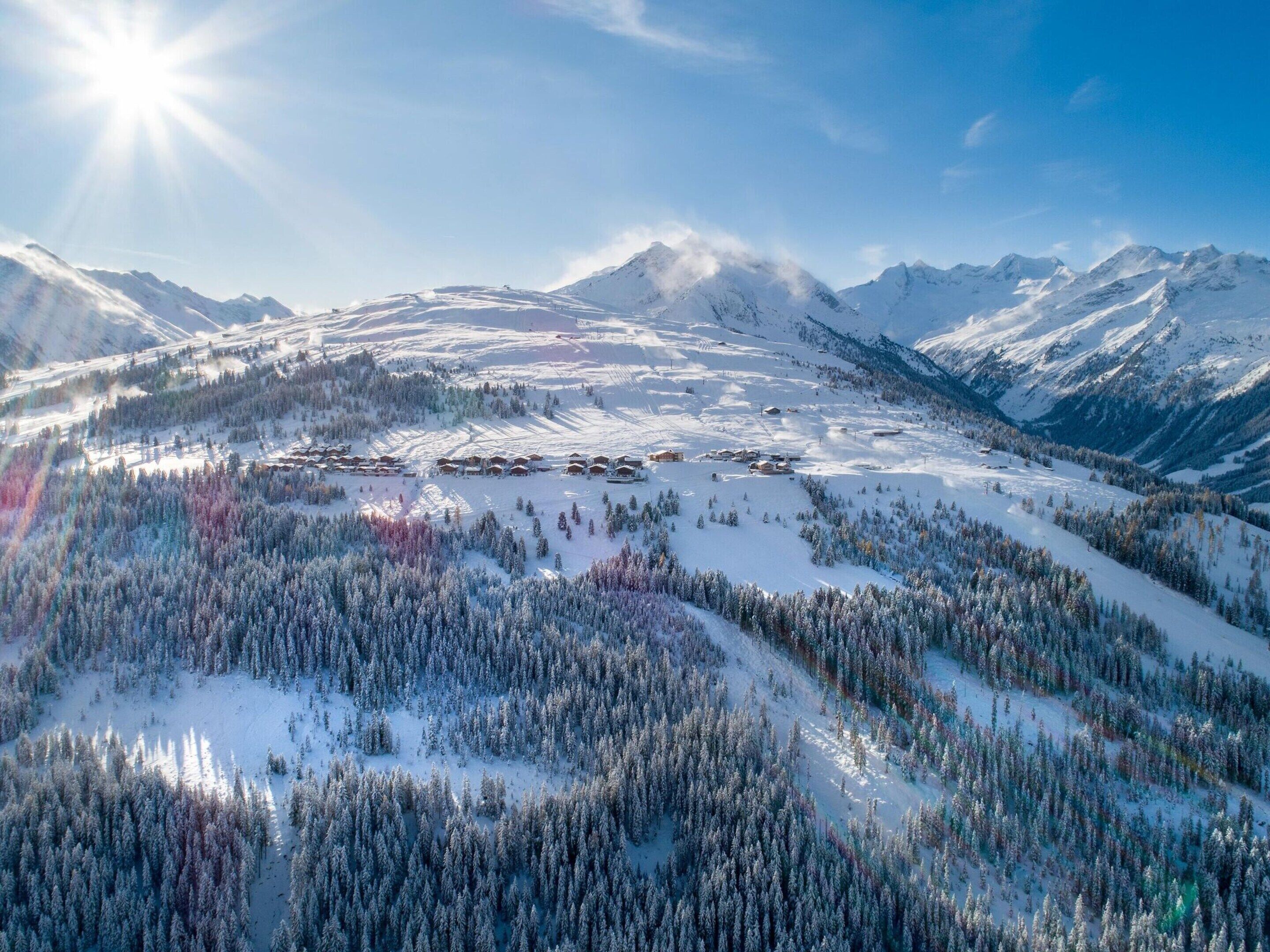 Paisagem montanhosa coberta de neve, com céu claro e sol brilhando à esquerda.
