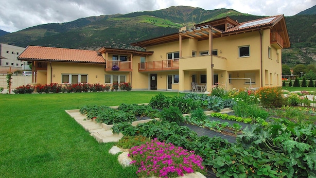 Um edifício de dois andares com varanda, rodeado por um jardim bem cuidado com diversas plantas e flores.