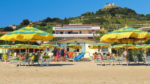 Uma praia com guarda-sóis listrados em amarelo e azul, piscina e um edifício com vista para uma colina.