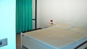 Quarto individual com uma cama branca, uma mesa de cabeceira com abajur vermelho, cortina verde e parede branca.