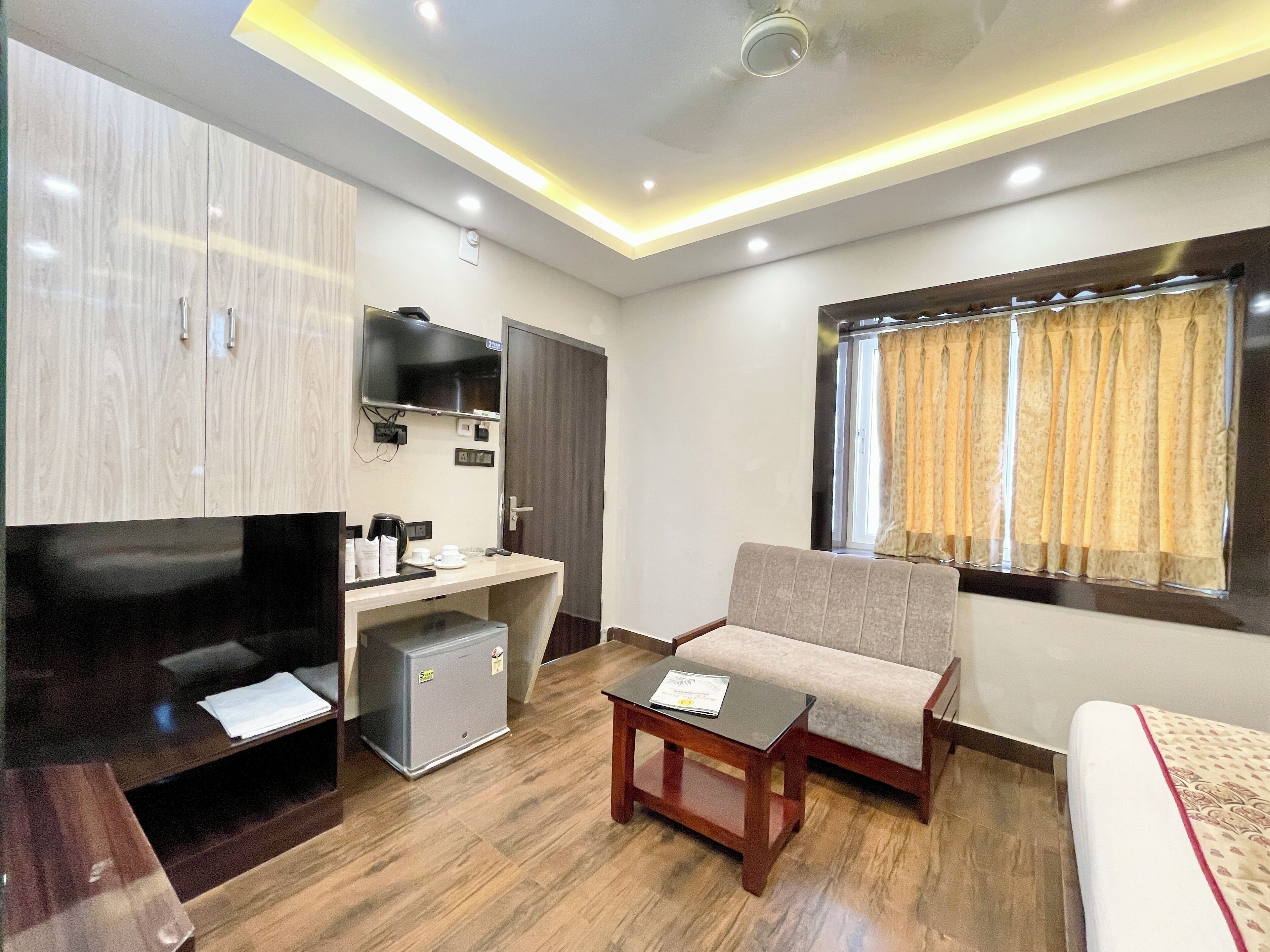 Quarto de hotel moderno com sofá, mesa pequena, televisão, escrivaninha com computador e janela com cortinas.