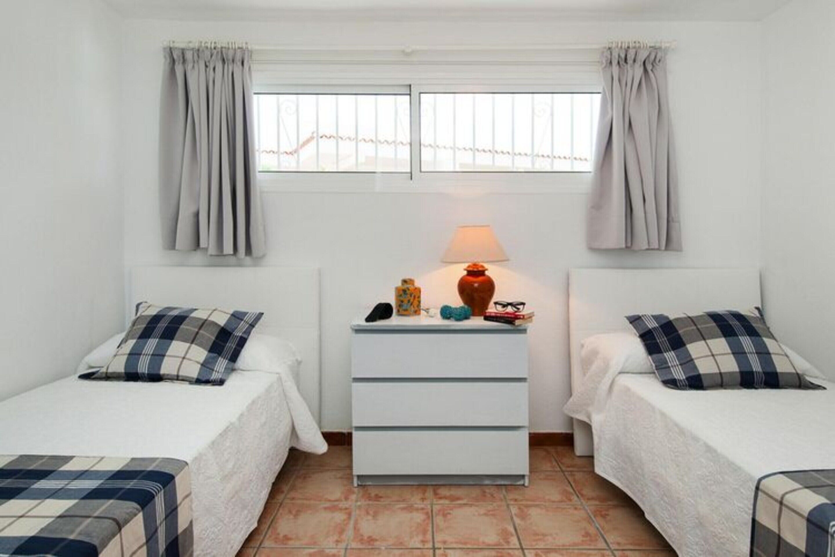 Quarto pequeno e limpo, com duas camas, uma mesinha de cabeceira, uma luminária e uma janela com cortinas.