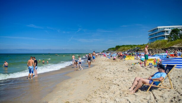 Uma praia com pessoas tomando sol e nadando, um edifício ao fundo e um céu azul límpido.