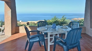 Mesa redonda com cadeiras e um vaso, disposta numa varanda com vista para o mar.