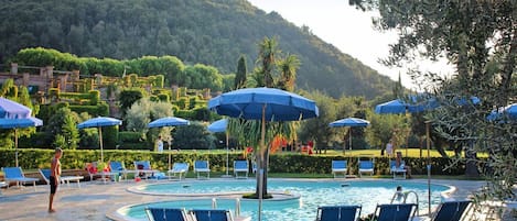 Área de piscina com espreguiçadeiras e guarda-sóis, rodeada por jardins bem cuidados e um horizonte montanhoso.