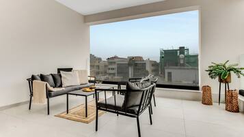 Uma sala de estar moderna com sofá, mesa de centro e cadeiras, uma janela grande com vista para a cidade e um vaso com planta.