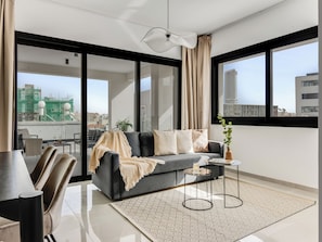 Uma sala de estar moderna com um sofá cinza, uma mesa de centro e uma vista para a cidade através de grandes janelas.