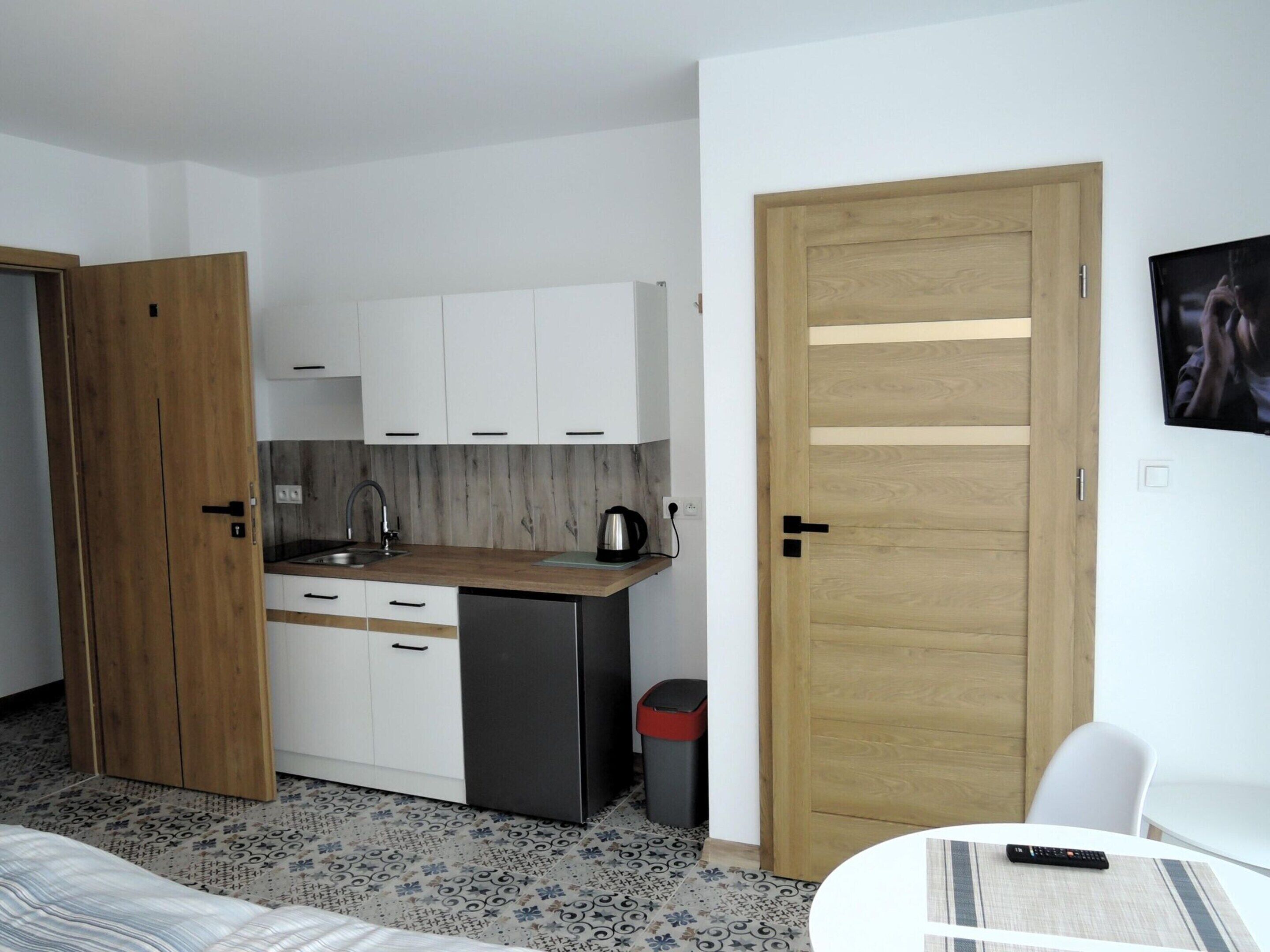 Cozinha compacta com armários brancos, pia e cafeteira. Há um cesto de lixo e uma porta que dá para outro quarto.