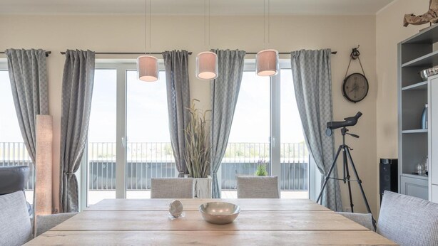 Uma área de refeições com uma mesa de madeira, cadeiras estofadas cinzentas e luminárias pendentes.