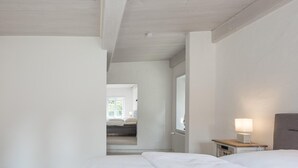 Quarto de hotel com uma cama grande, uma mesa de cabeceira com abajur e vista para outro quarto por uma porta aberta.
