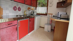 Uma cozinha compacta com armários vermelhos, uma pia de aço inoxidável, uma lava-louças branca e uma janela com cortina de renda.