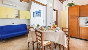 Uma cozinha compacta com armários de madeira, uma mesa de jantar posta para o café da manhã e um sofá azul em um quarto claro com piso de azulejos.