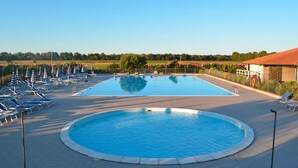 Uma piscina retangular ampla com uma área circular menor, rodeada por cadeiras de descanso e guarda-sóis, com vista desimpedida da paisagem ao redor.