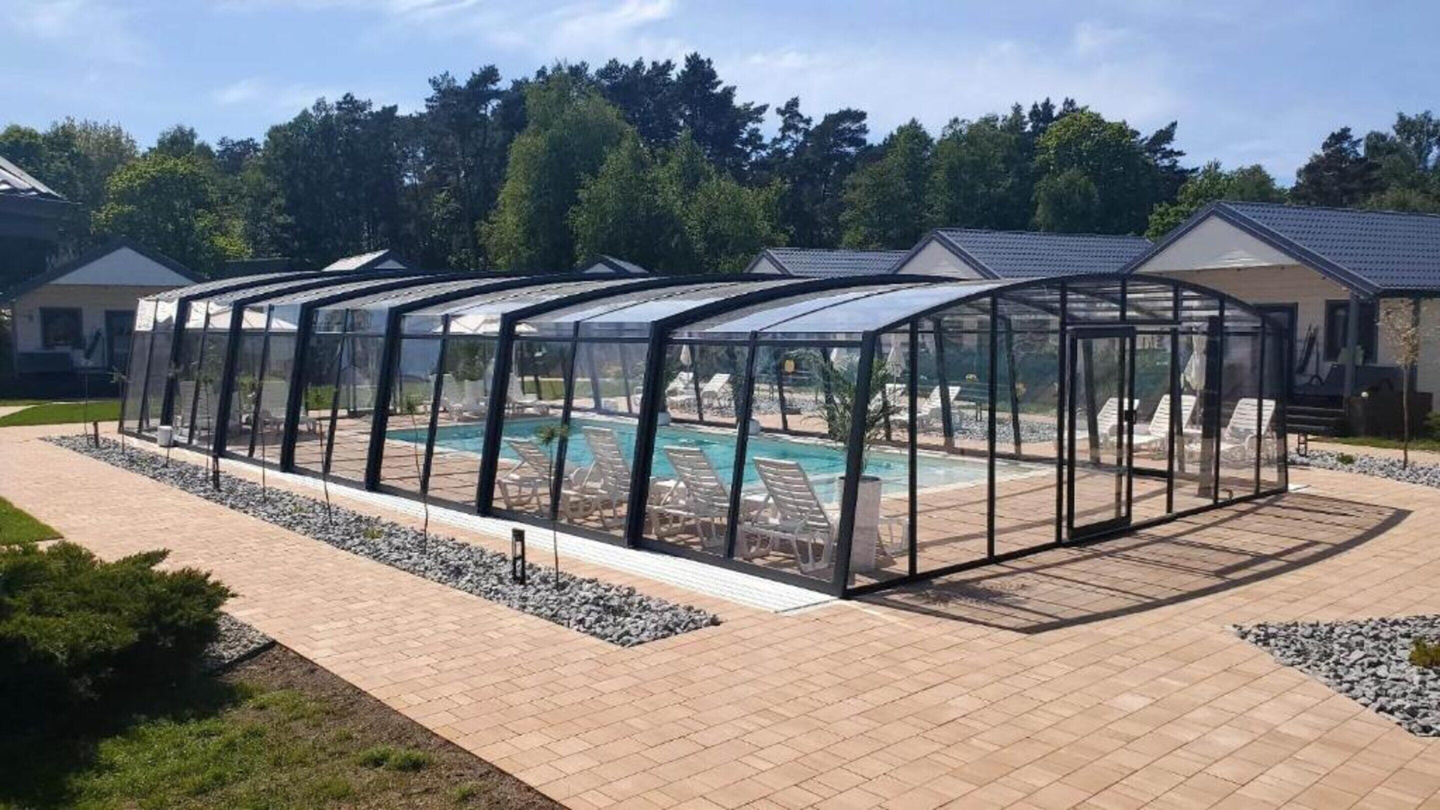 Piscina coberta ao ar livre com estrutura de vidro, rodeada por um pátio pavimentado e área de lazer com espreguiçadeiras.