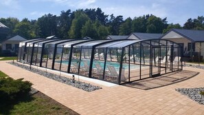 Piscina coberta ao ar livre com estrutura de vidro, rodeada por um pátio pavimentado e área de lazer com espreguiçadeiras.