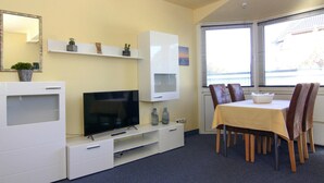 Uma sala de estar moderna com televisão de tela plana, armários brancos, uma mesa de jantar com cadeiras e uma janela grande com persianas.