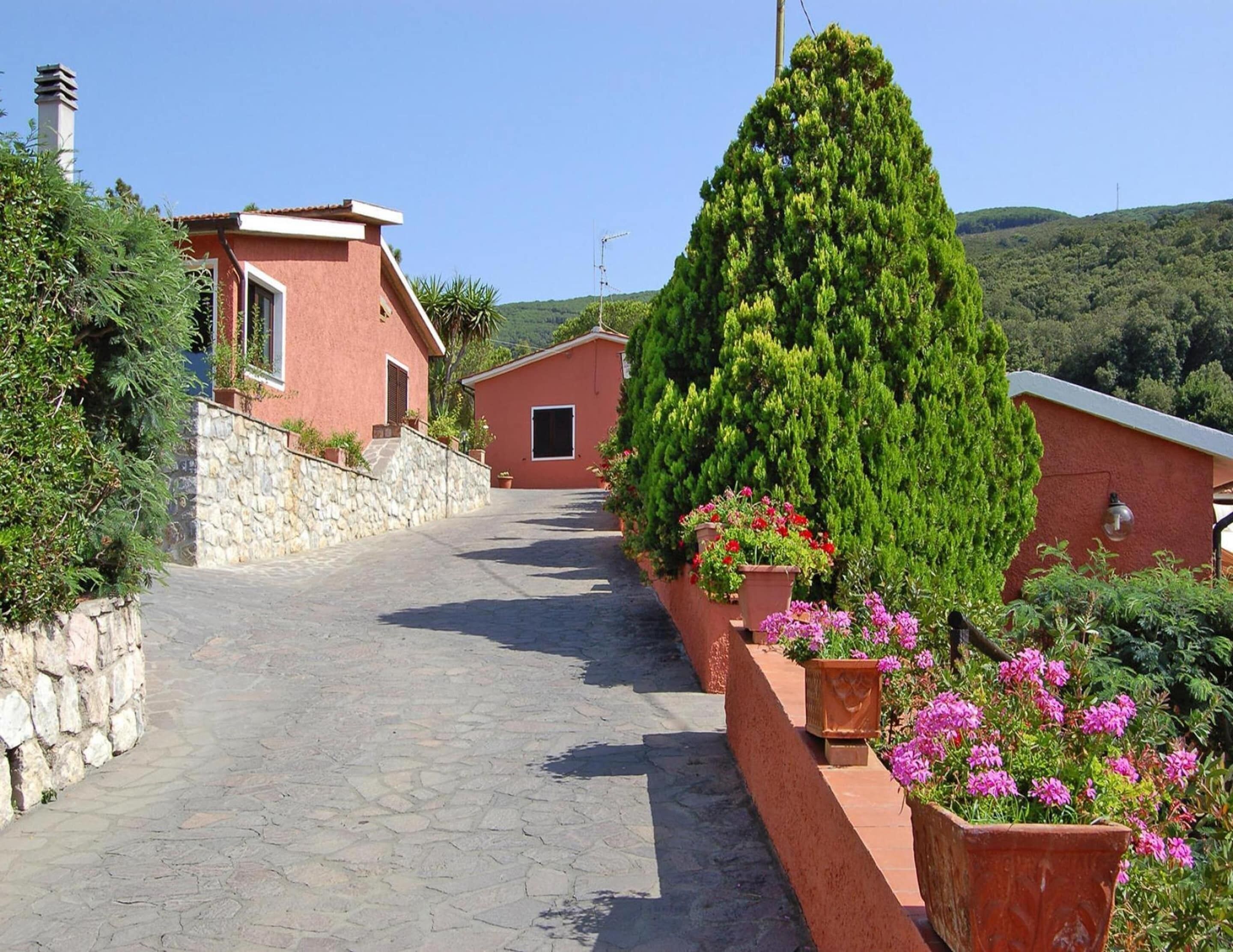 Um caminho estreito de pedras que leva a um edifício com telhado vermelho, com uma parede de pedra de um lado e uma fileira de vasos com flores rosa do outro.
