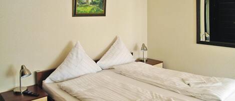 Uma cama de casal com roupa de cama branca, uma mesa de cabeceira com abajur e um quadro na parede.