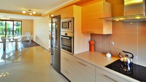 Cozinha moderna com eletrodomésticos de aço inoxidável, armários de madeira e área de jantar com mesa e cadeiras de vidro.