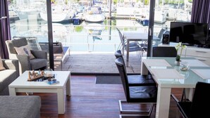 Sala de estar moderna com vista para uma marina. Inclui um sofá, uma mesa de centro, uma mesa de jantar e uma TV.