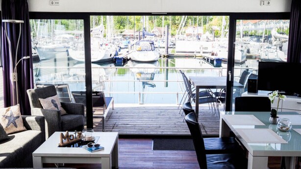 Quarto moderno com uma grande janela de vidro com vista para uma marina. Há um sofá, uma mesa de jantar e uma televisão.