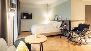 Quarto de hotel com cama, cadeira de rodas, mesa de cabeceira, abajur e televisão fixada na parede.