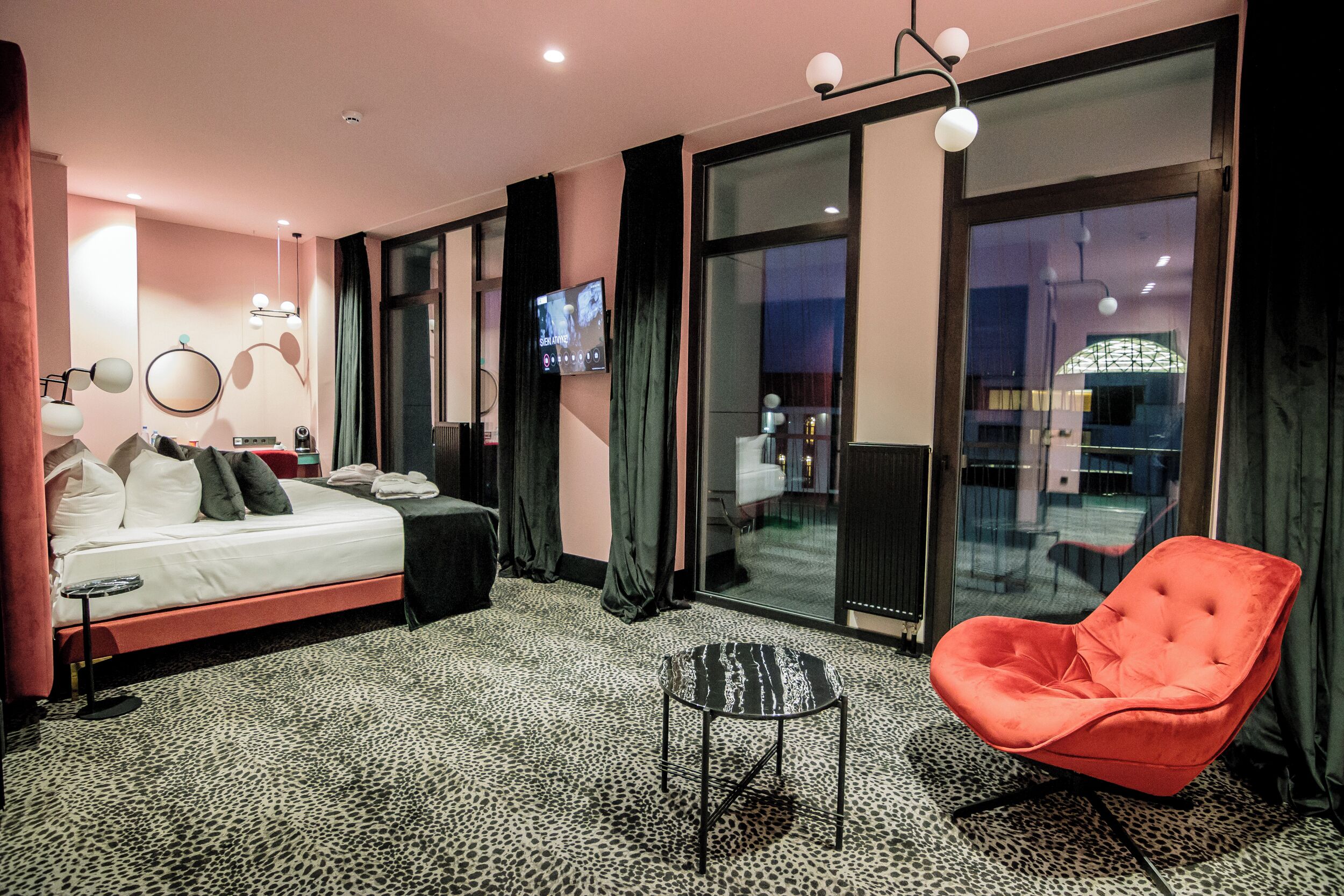 Quarto de hotel moderno com uma cama, uma poltrona vermelha, uma mesinha e uma televisão.