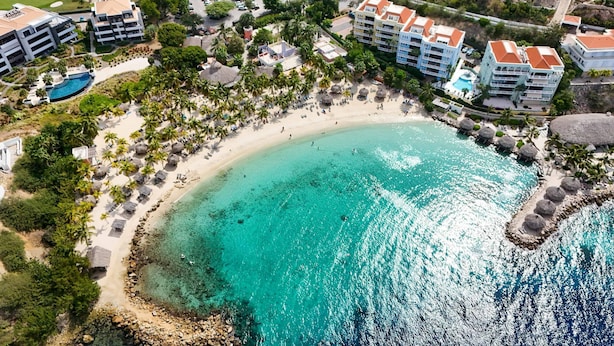 Vista aérea de um resort costeiro com praia de areia, águas turquesas cristalinas e vários edifícios do resort com varandas.