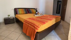 Cama bem arrumada com manta listrada, mesa de cabeceira com uma planta e um livro, e bandeja com uma garrafa e copos na cama.
