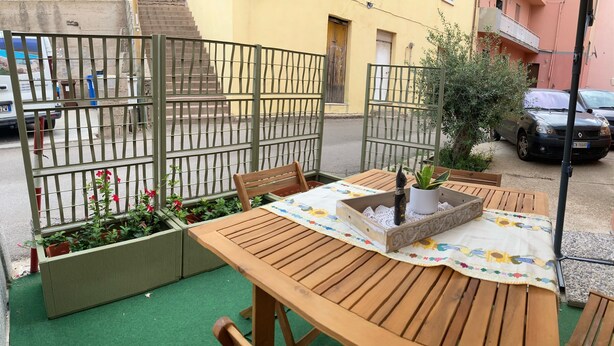 Uma mesa de madeira com toalha de mesa florida, rodeada por cadeiras, sobre uma superfície verde com plantas em vasos e uma caixa decorativa.