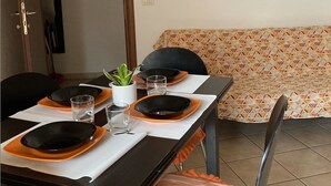 Uma área de refeições com uma mesa posta para dois, cadeiras, uma planta em vaso e um sofá estampado ao fundo.