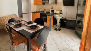 Cozinha compacta com geladeira, micro-ondas e fogão. Há uma mesa de jantar com cadeiras e um sofá ao fundo.