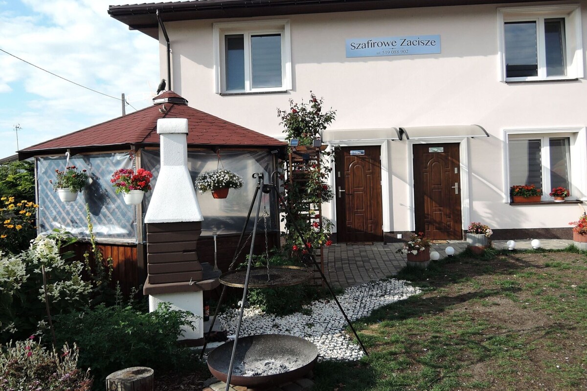 Uma casa de dois andares com uma placa indicando 'Szaffirowe Zaciszce' e um número de contato. Possui um pequeno jardim com um bebedouro para pássaros e plantas em vasos.