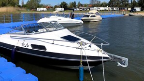 Uma marina com vários barcos atracados, um cais flutuante azul e uma fileira de casas ao fundo.