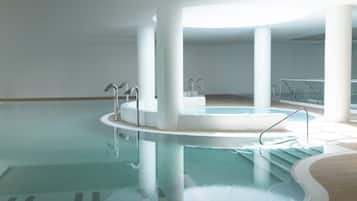 Uma grande piscina interior com água cristalina, rodeada por colunas brancas e um deck de madeira.