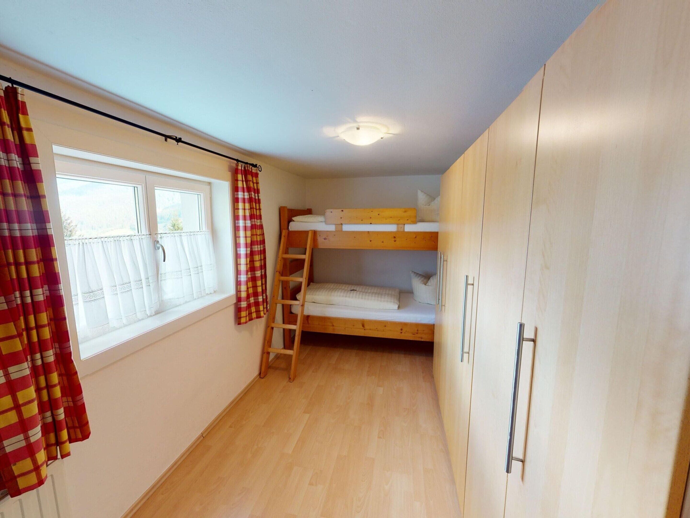 Um alojamento pequeno e individual, com uma cama beliche, uma escada de madeira, um guarda-roupa e uma janela com cortinas.