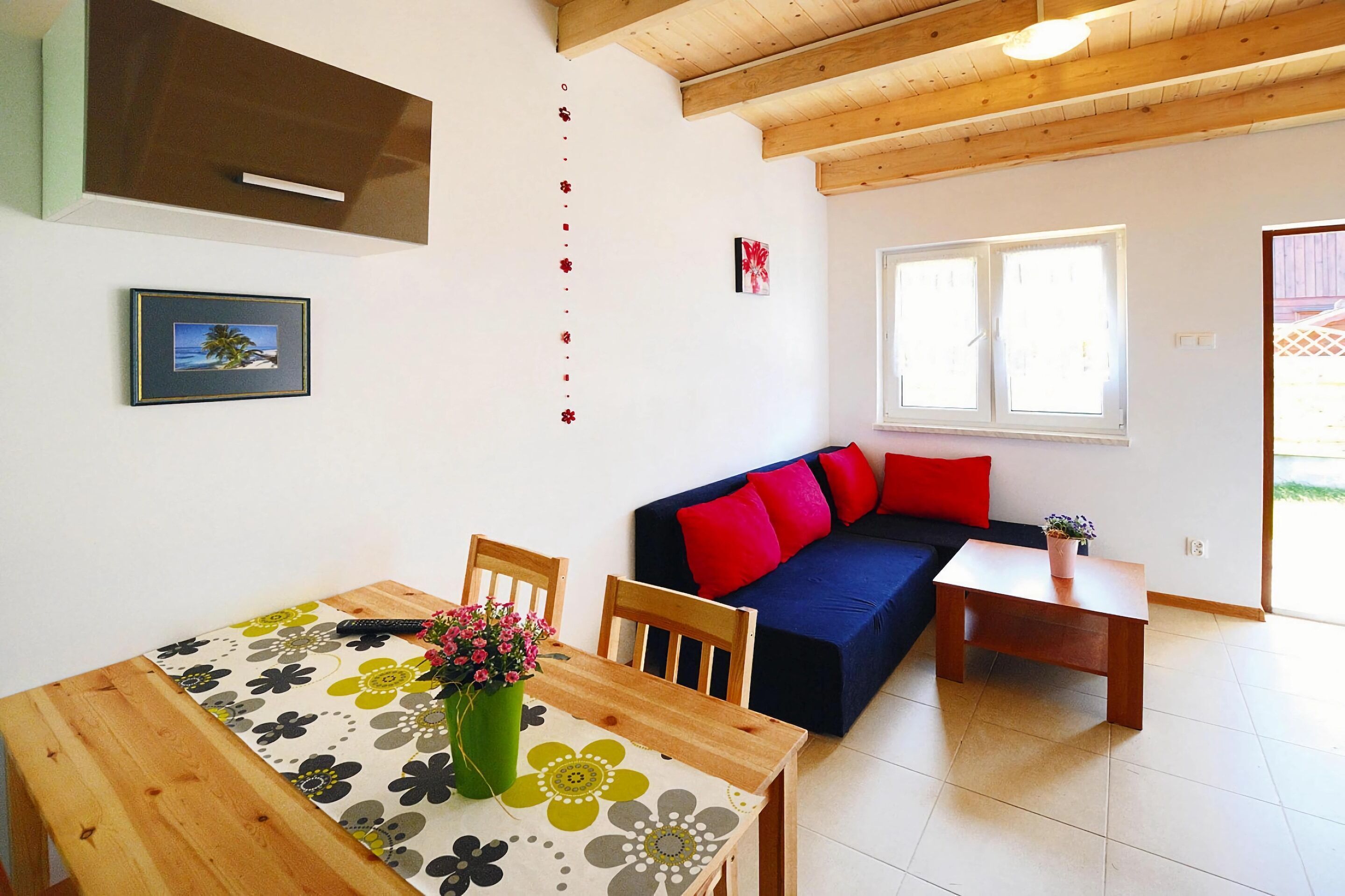 Um quarto com uma mesa de jantar de madeira, um sofá com almofadas vermelhas, uma pequena mesa com uma planta e um quadro na parede.