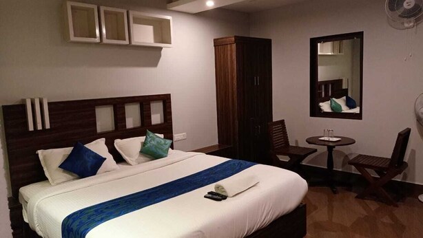 Quarto de hotel com uma cama grande, um guarda-roupa de madeira, uma pequena mesa redonda com uma cadeira e um ventilador fixado na parede.