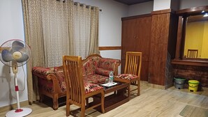 Uma sala de estar com um sofá, duas cadeiras de madeira, uma mesinha com uma garrafa de água, um ventilador de chão e um guarda-roupa de madeira.