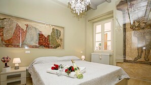 Quarto de hotel com uma cama grande, mesa de cabeceira com abajur, janela com vista e um candelabro.