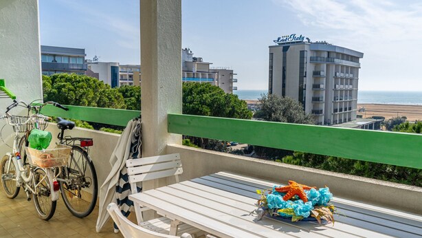 Uma varanda com mesa e cadeiras brancas, uma bicicleta e vista para a praia, com o Hotel Italia ao fundo.