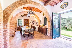Cozinha rústica com arcos de tijolos aparentes, mesa de jantar posta para quatro pessoas e vista do jardim através de uma porta aberta.