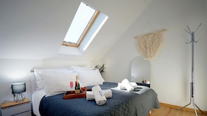 Quarto com claraboia, cama com colcha escura, mesa de cabeceira com abajur, espelho e luminária de chão.