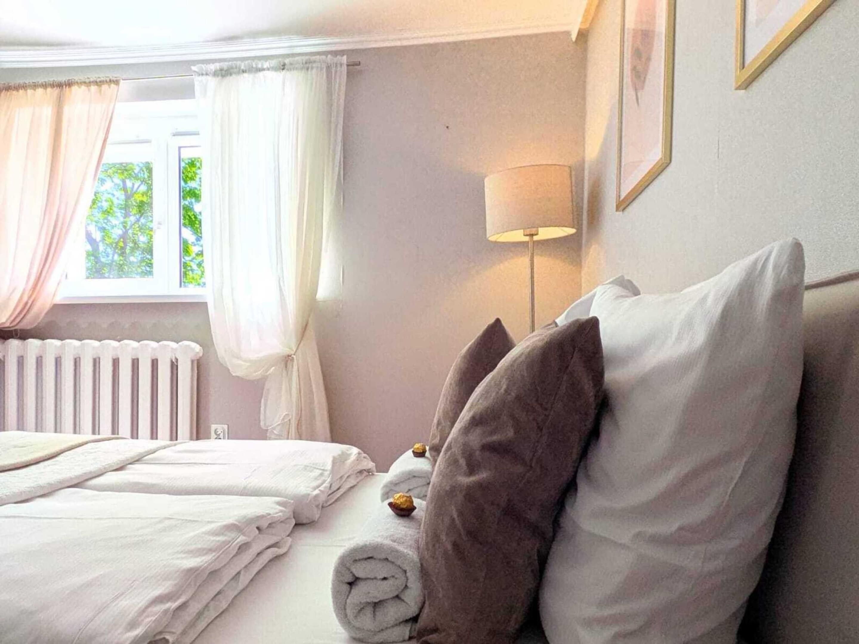 Cama bem arrumada com lençóis brancos e duas almofadas, um radiador, uma janela com cortinas e uma luminária na mesa de cabeceira.