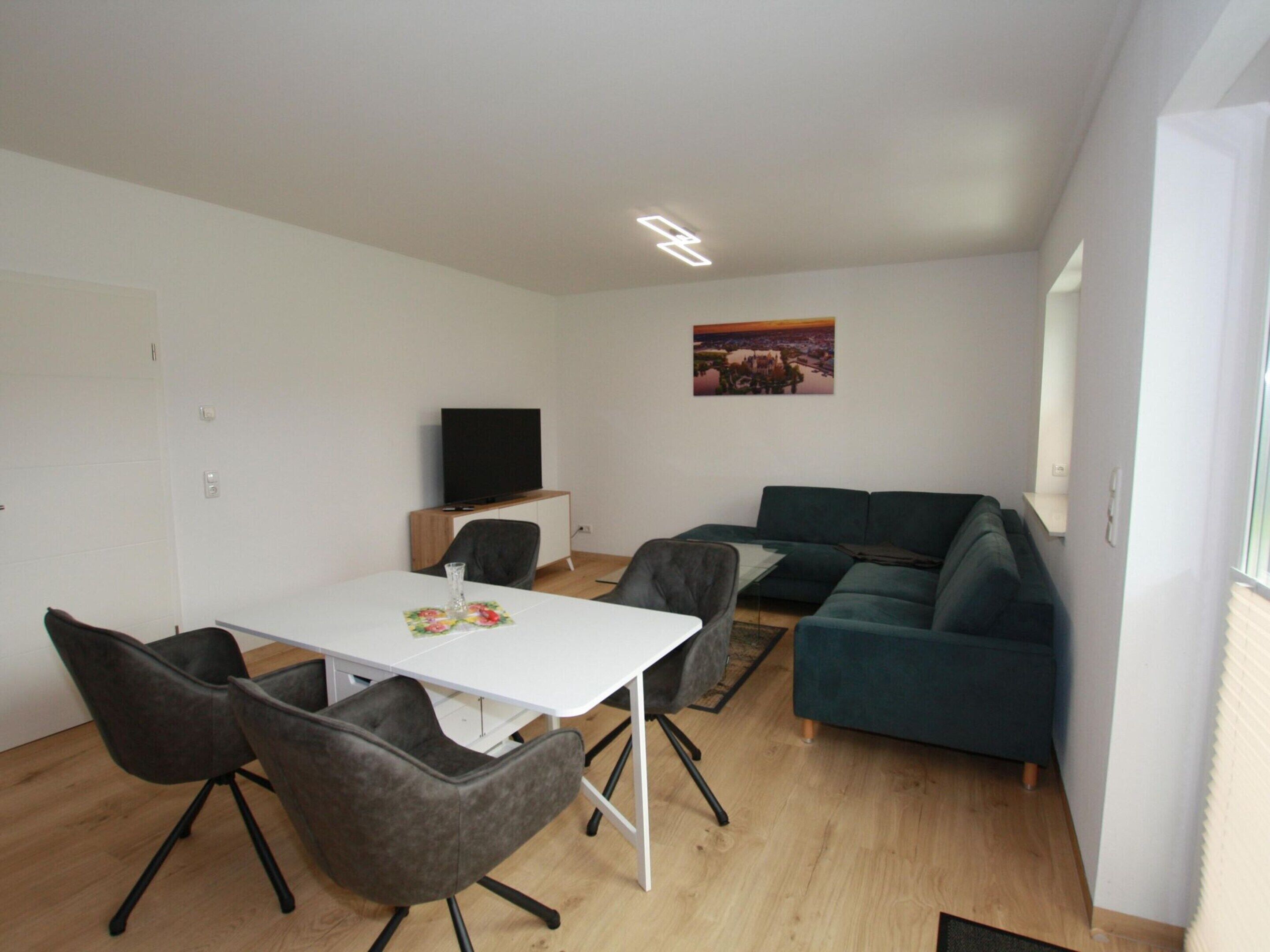 Uma sala de estar moderna com uma mesa branca, duas cadeiras cinzas, um sofá verde-água e uma TV de tela plana em um suporte de madeira.