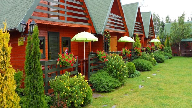 Uma fileira de cabanas de madeira com telhados verdes, cada uma com uma varanda e um pequeno jardim com flores e arbustos.