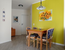 Uma sala de jantar com uma mesa e cadeiras de madeira, um quadro de girassóis na parede e uma porta ao fundo.