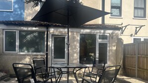 Um pátio com uma mesa e quatro cadeiras, um guarda-sol grande e vista para uma casa com janela e porta.