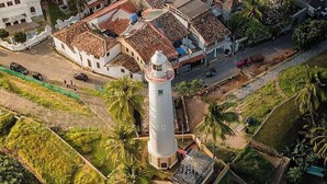 Vista aérea de uma cidade costeira com um farol branco imponente, rodeada por edifícios residenciais, uma praia e palmeiras.