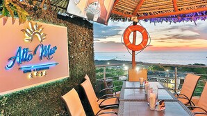 Restaurante com mesas ao ar livre e vista para o mar, com um letreiro de neon, um salva-vidas e um telhado de palha.